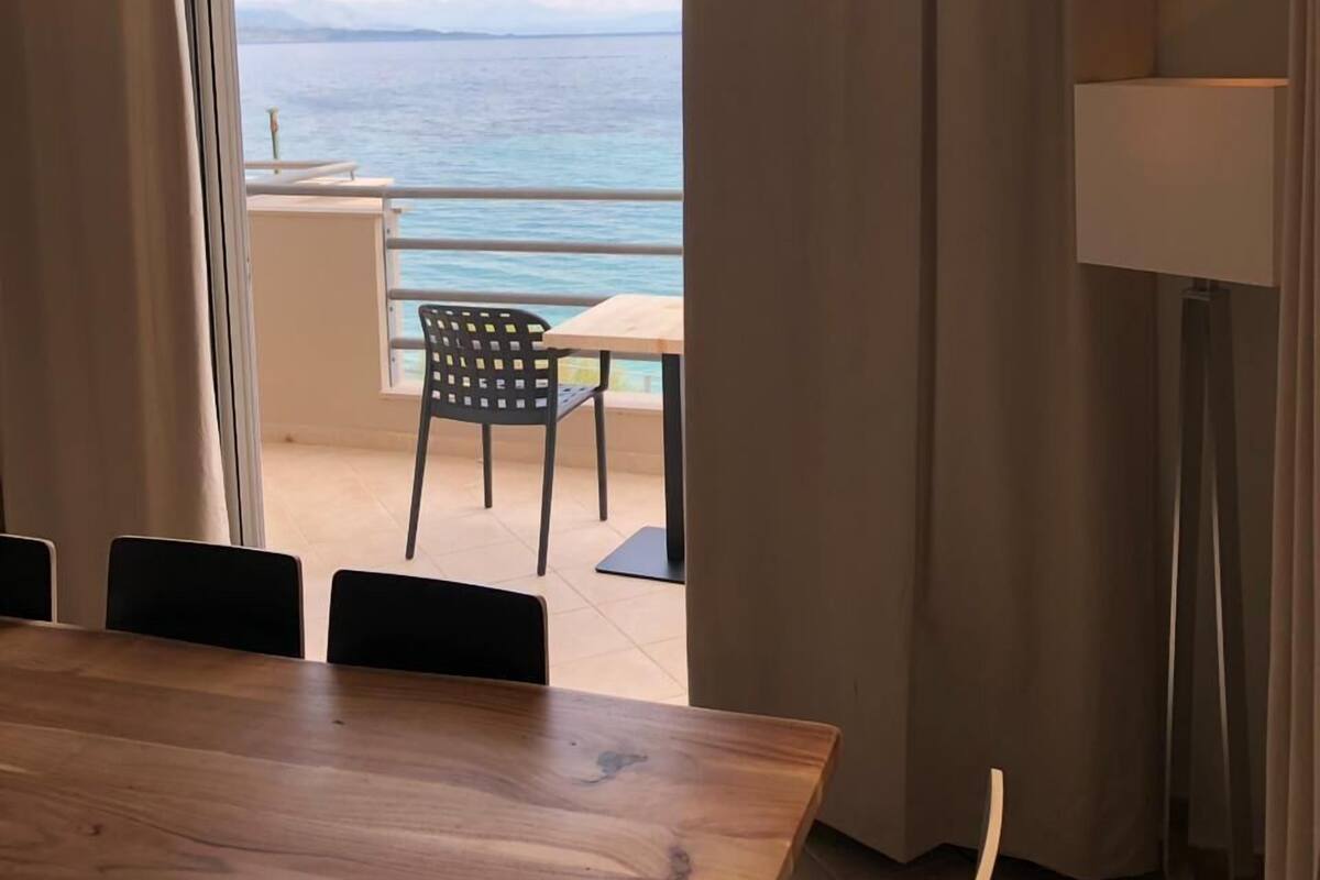 Um quarto com mesa e cadeiras de madeira, vista para o mar por uma grande janela e uma varanda com mesa e cadeiras.