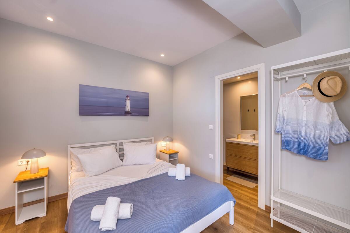 Quarto com cama, duas mesas de cabeceira com luminárias, pintura de um farol, guarda-roupa e banheiro visível por uma porta aberta.