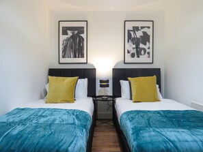 Quarto de hotel com duas camas, cada uma com colcha azul e travesseiro amarelo. Há quadros nas paredes acima das camas.