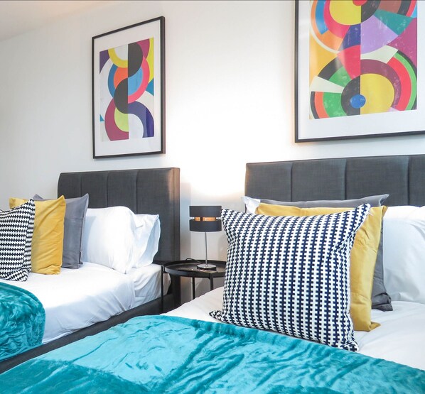 Quarto de hotel com duas camas, cada uma com cabeceira cinza, roupa de cama branca e almofadas estampadas. Há uma mesa pequena com luminária e arte abstrata nas paredes.