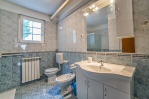 Casa de banho com sanita, bidé e lavatório, com azulejos em tons claros e uma janela sobre a sanita.