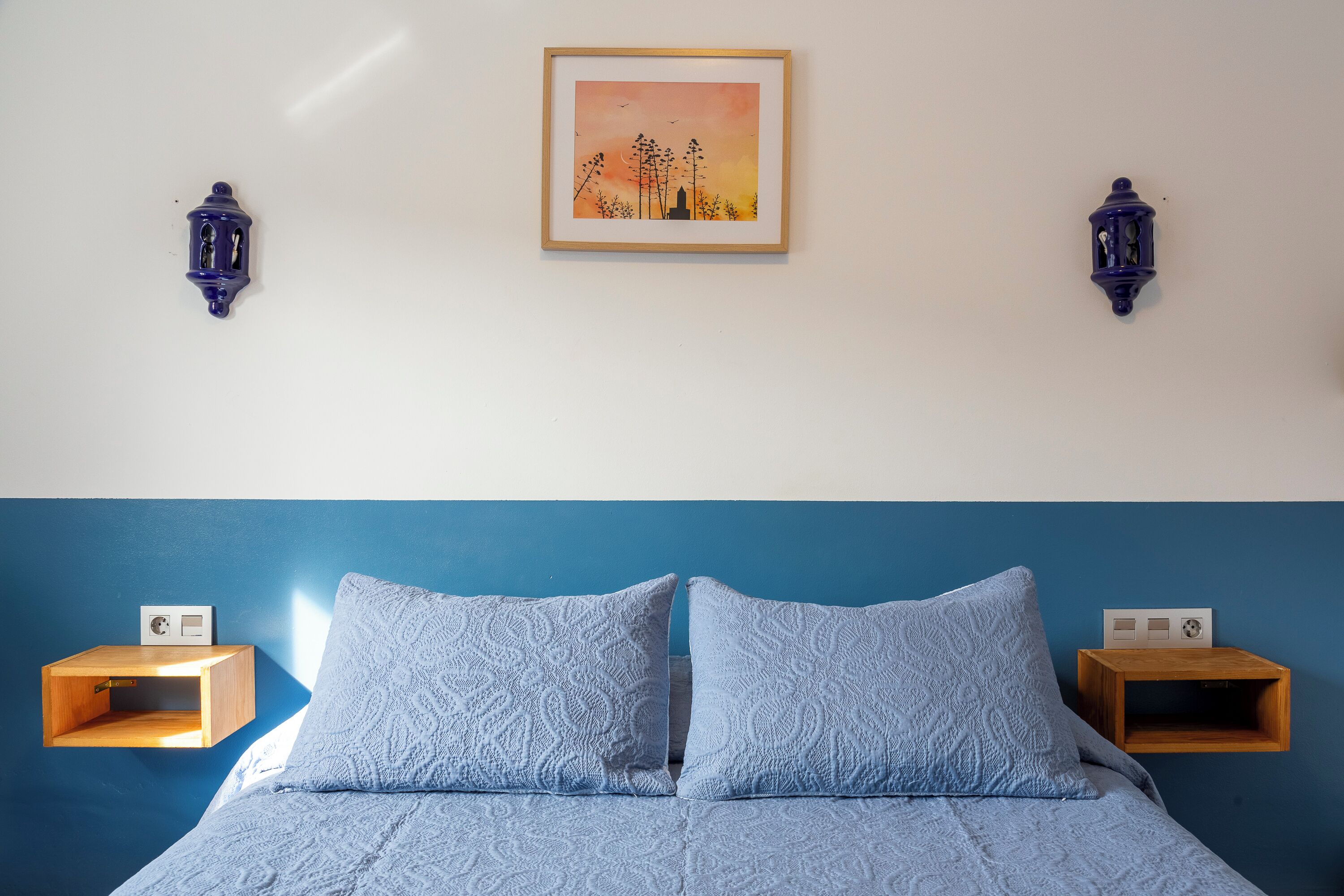 Quarto com cama, duas mesas de cabeceira fixadas na parede e um quadro na parede.