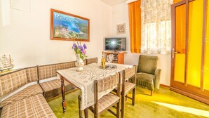 Uma área de refeições com uma mesa posta para dois, um espaço de bancos, uma televisão e uma porta com cortinas.