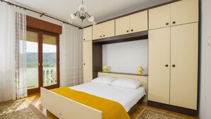 Quarto com cama, mesa de cabeceira, janela com cortinas e uma luminária no teto, com vista para o verde lá fora.
