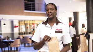 Uma pessoa vestindo uniforme branco e avental marrom, segurando um saco de papel marrom em um ambiente interno bem iluminado, com mesas e cadeiras.