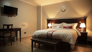 Quarto de hotel com cama, luminárias de cabeceira, televisão presa à parede, um banco de madeira e uma escrivaninha com cadeira.