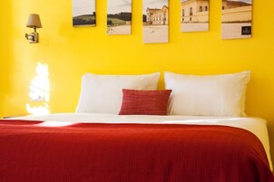 Quarto de hotel com parede amarelo vibrante, cama com roupa de cama branca e vermelha, e quatro quadros na parede.