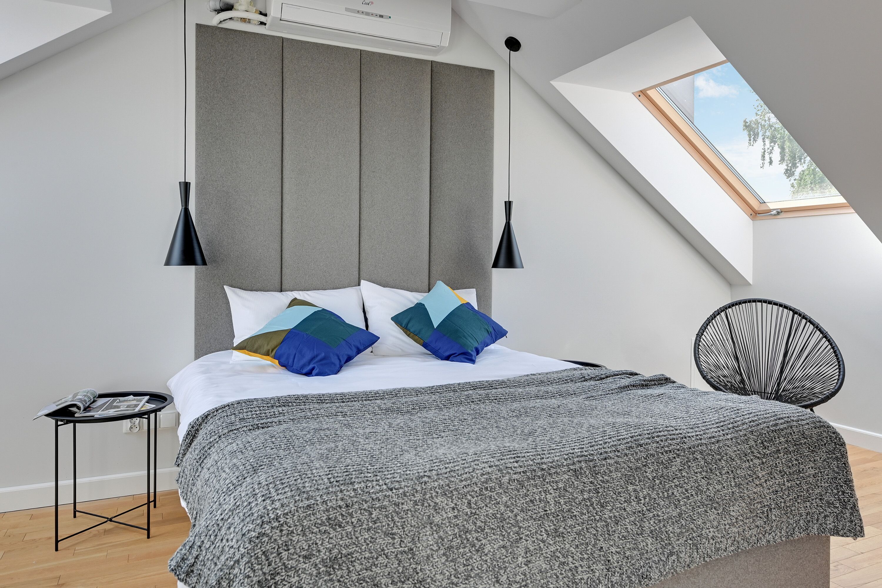 Quarto moderno com teto inclinado, claraboia, cama com manta cinza e duas luminárias pendentes pretas.