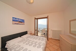 Quarto com cama, cômoda, radiador, pintura e varanda com vista.