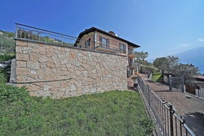 Um edifício de pedra com varanda, uma parede de pedra e um espaço pavimentado com guarda-corpo de metal.