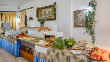 Disposição de buffet com diversos alimentos, um quadro de menu e elementos decorativos como uma pintura e uma planta.
