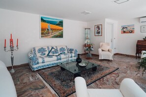 Uma sala de estar com sofá, poltronas, mesa de centro e tapete. Há quadros nas paredes e um candelabro no chão.