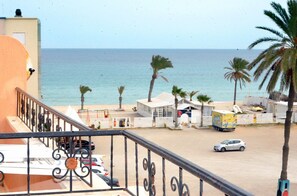 Vista à beira-mar com varanda, palmeiras, cabanas de praia e um carro estacionado.