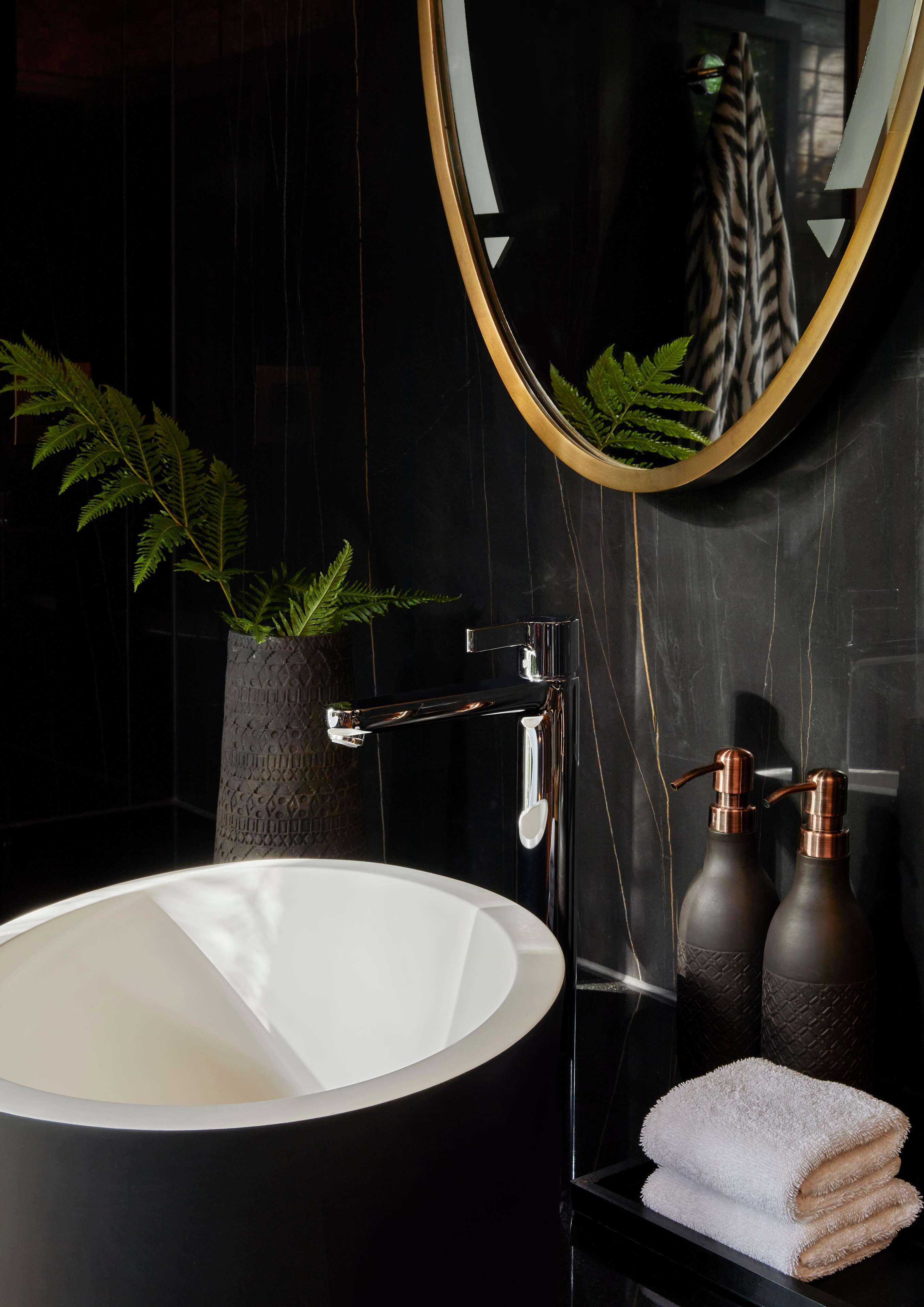 Casa de banho moderna com lavatório preto, uma sanita branca, espelho com moldura dourada e uma planta num vaso texturizado.