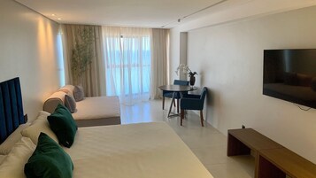 Quarto de hotel com cama, escrivaninha, cadeira, televisão e uma janela grande com cortinas.