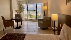 Quarto de hotel com cama, escrivaninha com cadeira, mesinha de centro com abajur e janela com vista para a praia.