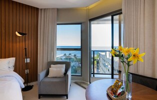 Quarto de hotel com cama, sofá, mesa de jantar, um vaso com flores e vista para o mar.