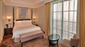 Quarto de hotel com uma cama grande, um banco, uma mesa redonda e uma cadeira. Há uma janela ampla com cortinas, um quadro na parede e uma mesa de cabeceira com uma luminária.