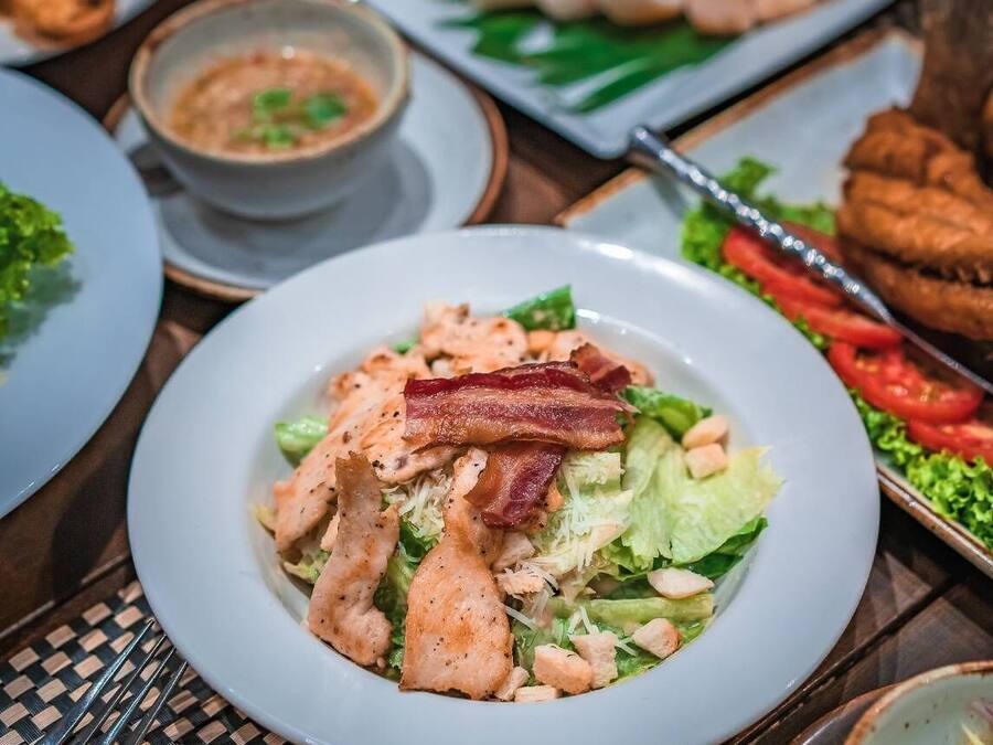 Uma mesa com diversos pratos, incluindo uma salada com bacon, frango grelhado e outros alimentos.