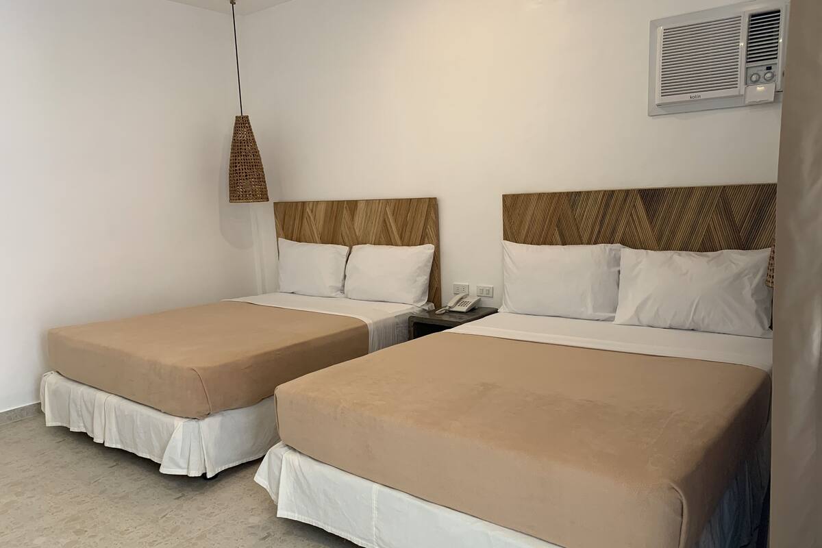 Quarto de hotel com duas camas, cada uma com cabeceira e roupa de cama branca. Há um telefone na mesa de cabeceira e um aparelho de ar condicionado fixado na parede.