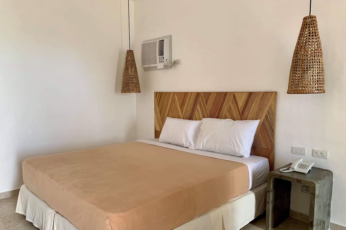 Quarto com cama grande, cabeceira de madeira, duas luminárias de vime penduradas, telefone na mesa de cabeceira e ar condicionado.