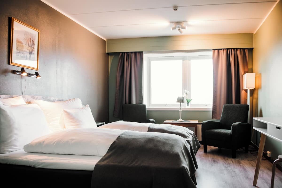 Quarto de hotel com uma cama grande, duas mesas de cabeceira, uma cadeira, uma escrivaninha e uma janela com cortinas.