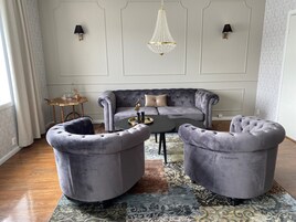 Uma sala de estar com sofá cinzento, duas poltronas, uma mesa de centro e um candelabro.
