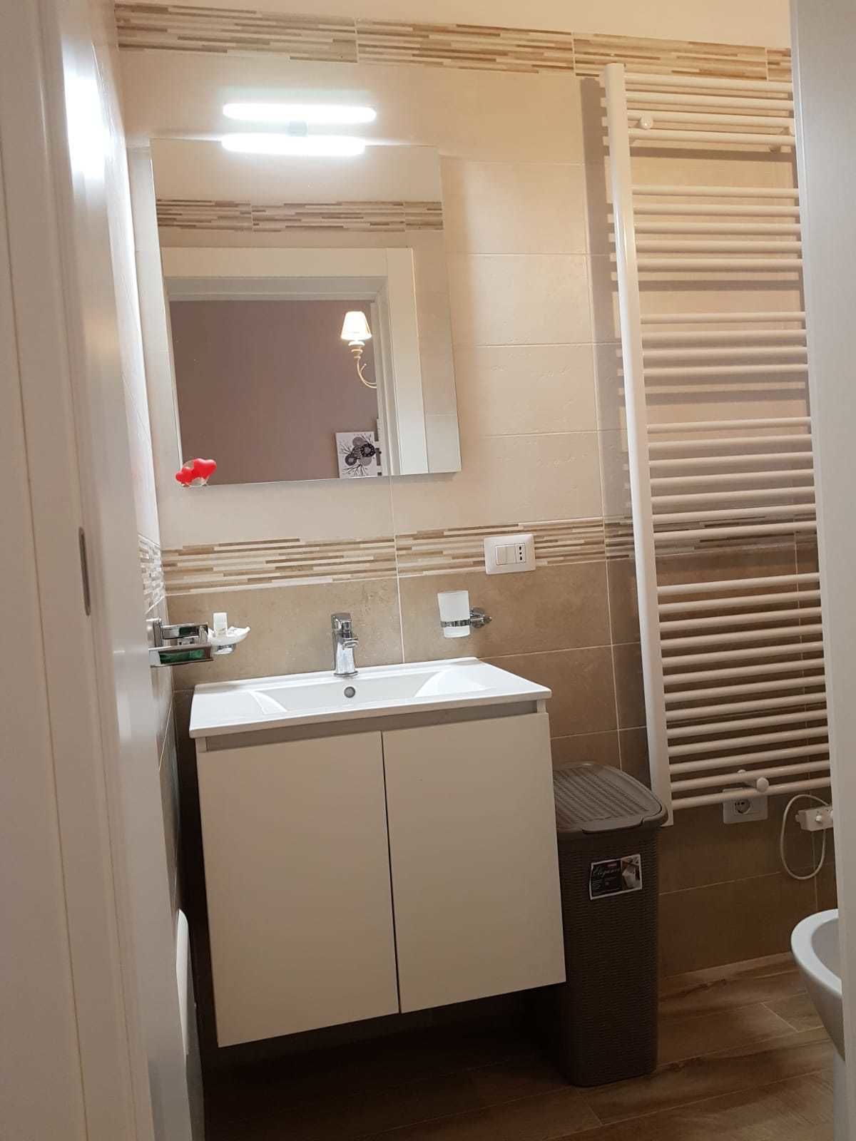 Uma casa de banho com uma pia branca, espelho e um porta-toalhas. Há um cesto de lixo e um dispensador de toalhas na parede.