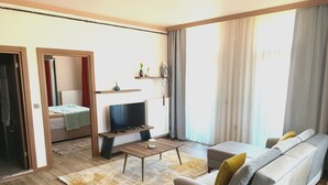 Uma sala de estar com um sofá, uma mesa de centro e uma televisão de tela plana fixada na parede. Há uma janela com cortinas, uma porta que dá para outro cômodo e um pequeno aparador com objetos decorativos.
