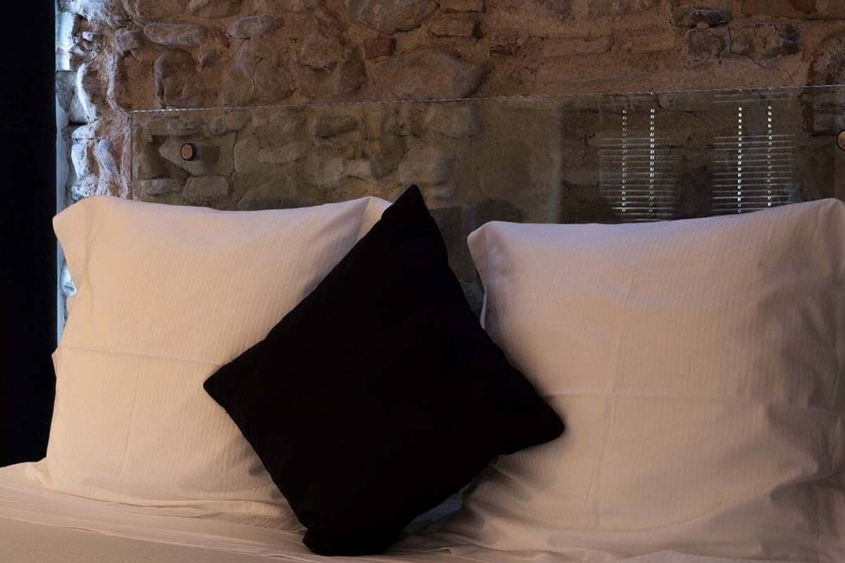 Cama com roupa de cama branca e almofada preta em quarto com parede de pedra e cabeceira de vidro.