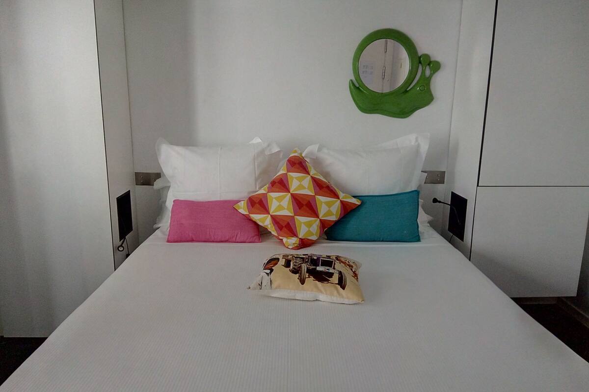 Uma cama com lençóis brancos, um espelho verde fixado na parede e três almofadas em rosa, vermelho e azul.