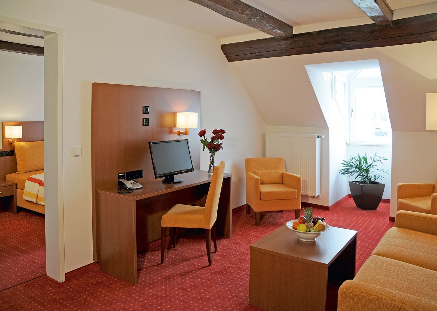 Quarto de hotel moderno com uma escrivaninha de madeira, um computador, uma cadeira, um sofá e uma pequena mesa com frutas. Há uma planta em vaso e um radiador.