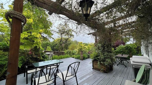 Um deck de madeira com mesa e cadeiras, cercado por vegetação exuberante e uma pérgola com luzes penduradas.