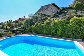 Uma piscina com água azul cristalina, rodeada por sebes bem cuidadas e um edifício de pedra ao fundo.
