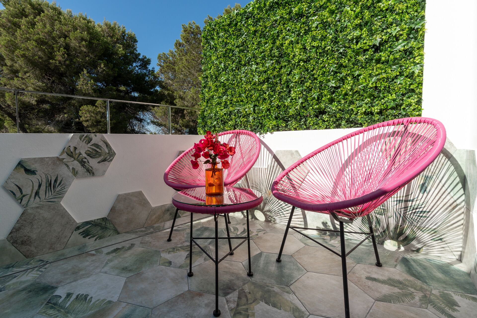 Um pátio com duas cadeiras de vime cor-de-rosa, uma mesa pequena e um vaso com flores vermelhas.