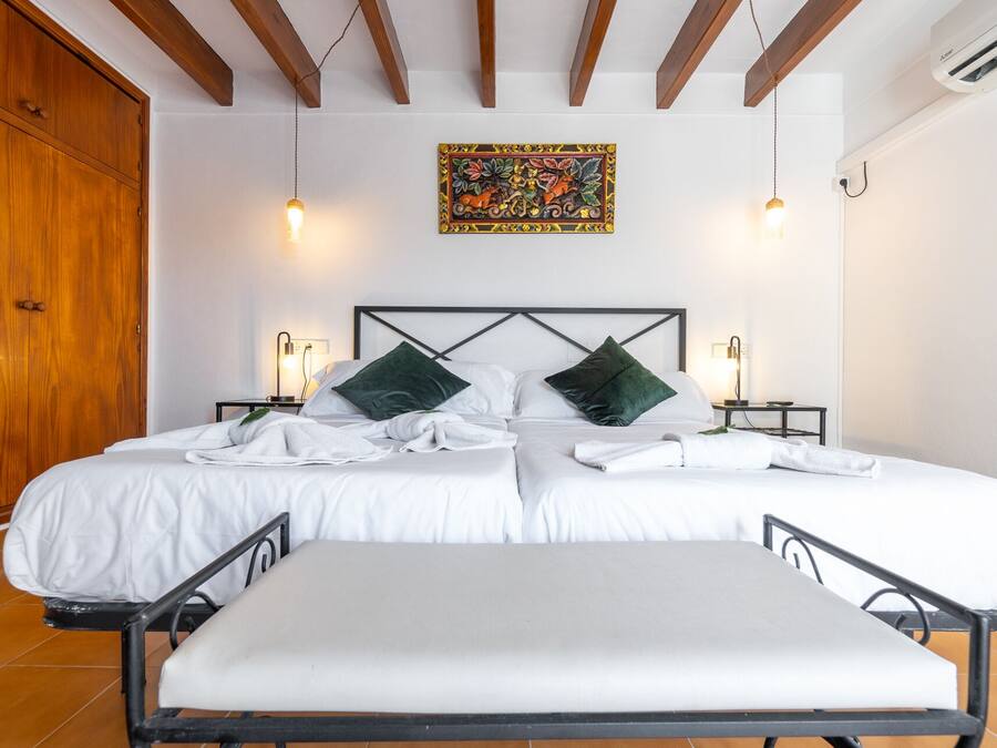 Uma cama de casal com roupa de cama branca, duas mesas de cabeceira com luminárias, teto de madeira e um adorno colorido na parede.