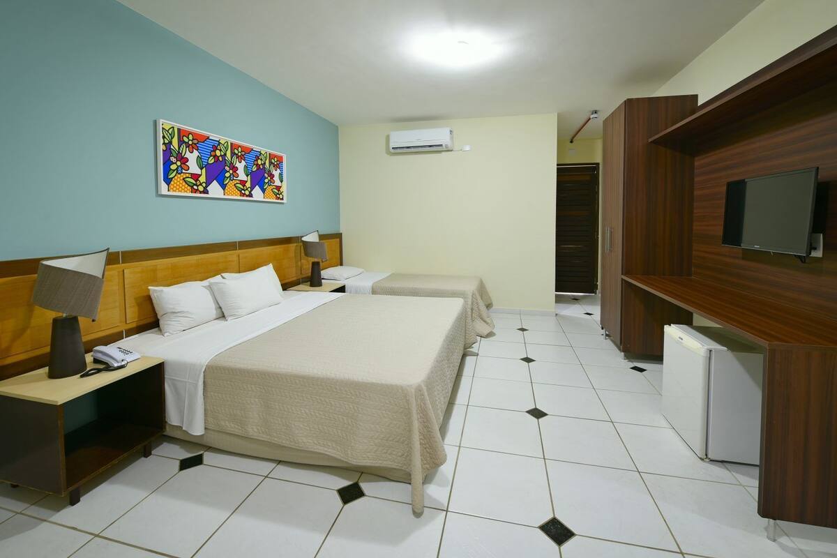 Quarto de hotel com uma cama grande, mesas de cabeceira, televisão, ar condicionado e um quadro colorido na parede.