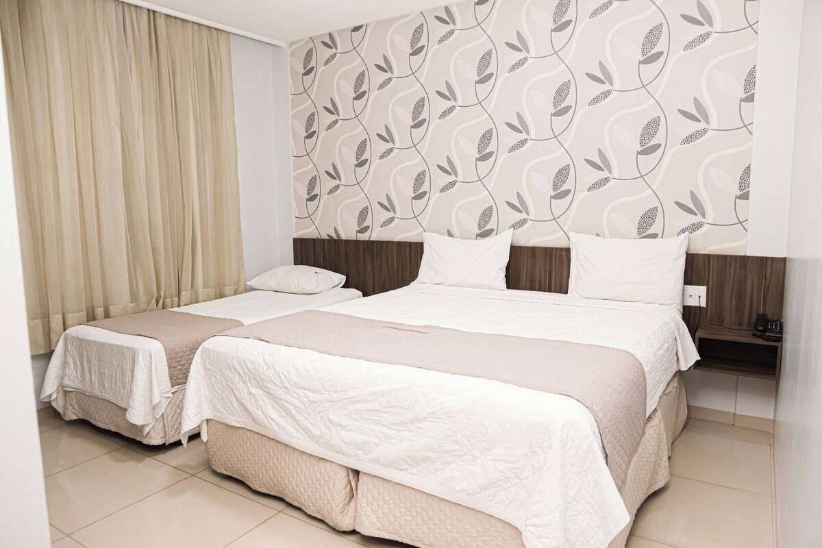 Quarto de hotel com duas camas, uma janela grande com cortinas, parede estampada e mesa de cabeceira com telefone.