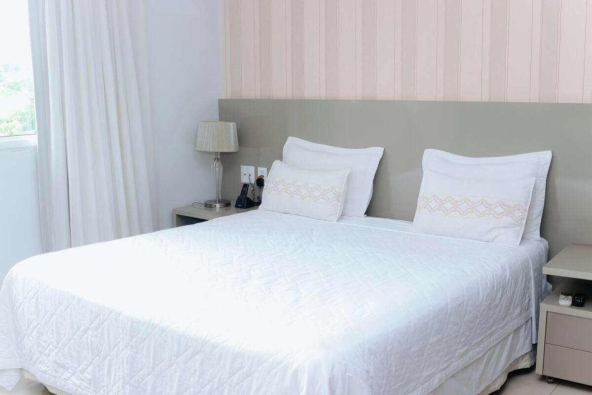 Uma cama bem arrumada com um edredom branco, dois travesseiros e uma mesa de cabeceira com abajur e telefone.