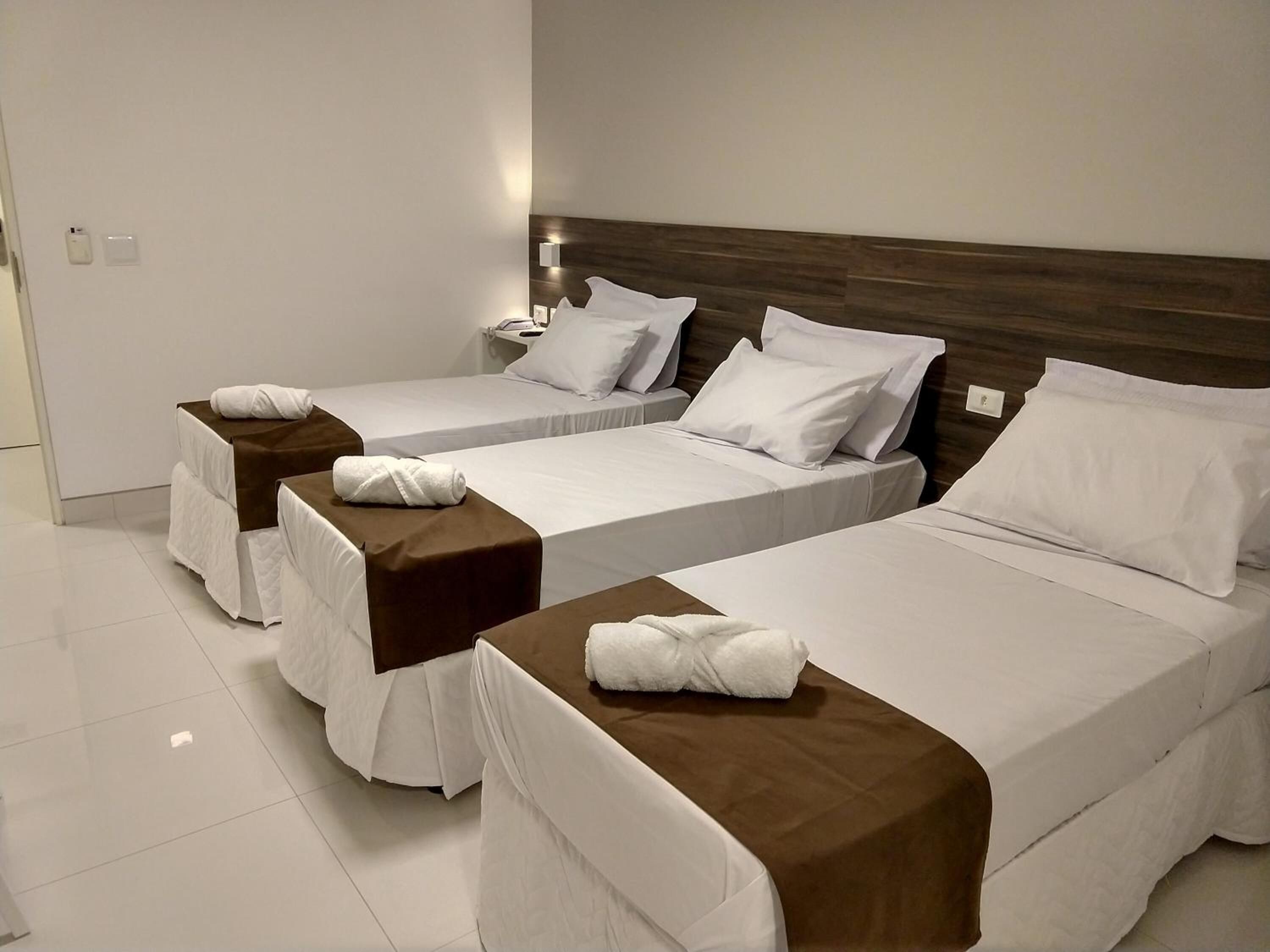 Quarto de hotel com três camas de solteiro, cada uma com cabeceira marrom e roupa de cama branca.