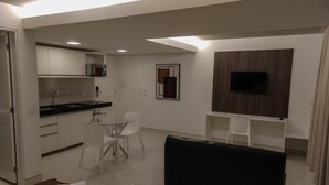 Sala de estar moderna com um sofá preto, uma mesa de centro de vidro e duas cadeiras brancas. Há uma área de cozinha com armários brancos e micro-ondas. Uma televisão de tela plana está instalada em uma parede revestida de madeira.