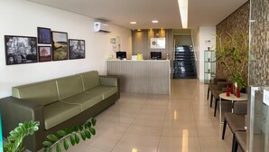 Um hall de hotel moderno com uma mesa de recepção, um sofá verde, uma área de refeições e uma escada.