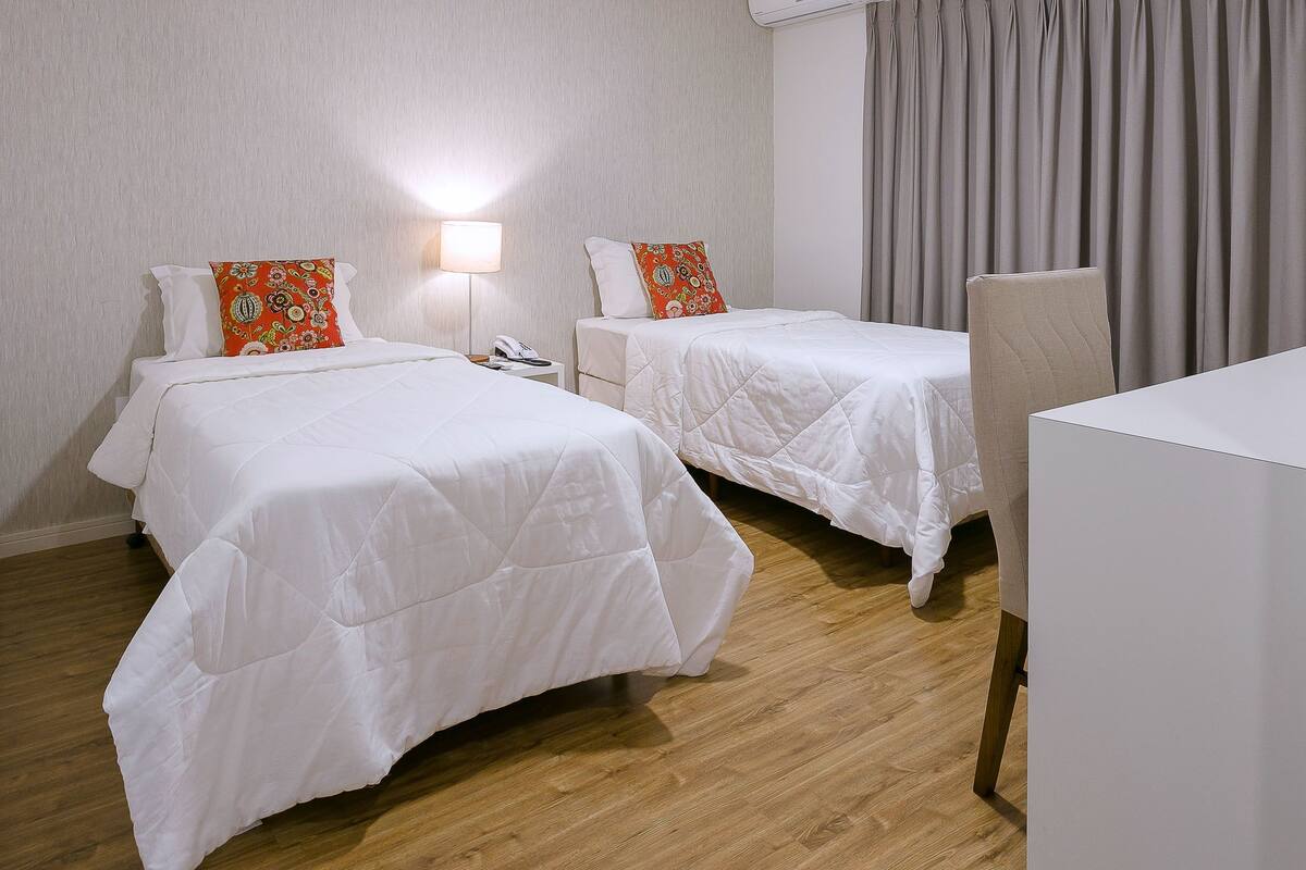 Quarto de hotel com duas camas, uma mesinha de cabeceira com abajur e uma cadeira.