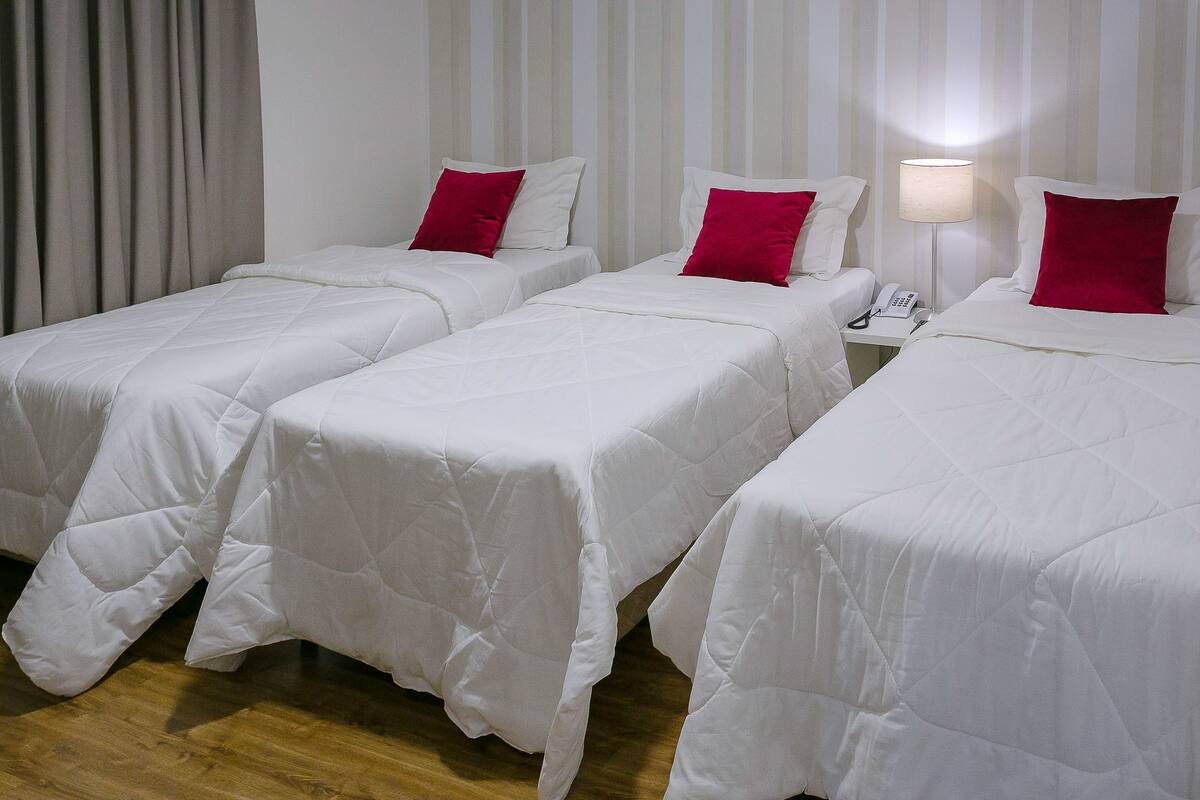 Quarto de hotel com duas camas de solteiro, cada uma com roupa de cama branca e travesseiro vermelho. Há uma mesinha de cabeceira com abajur e telefone.
