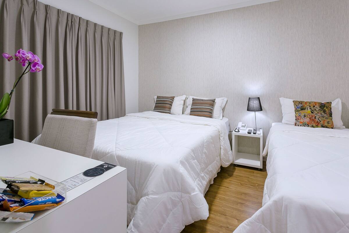 Quarto de hotel com duas camas, uma escrivaninha com petiscos, uma luminária e um travesseiro decorativo.