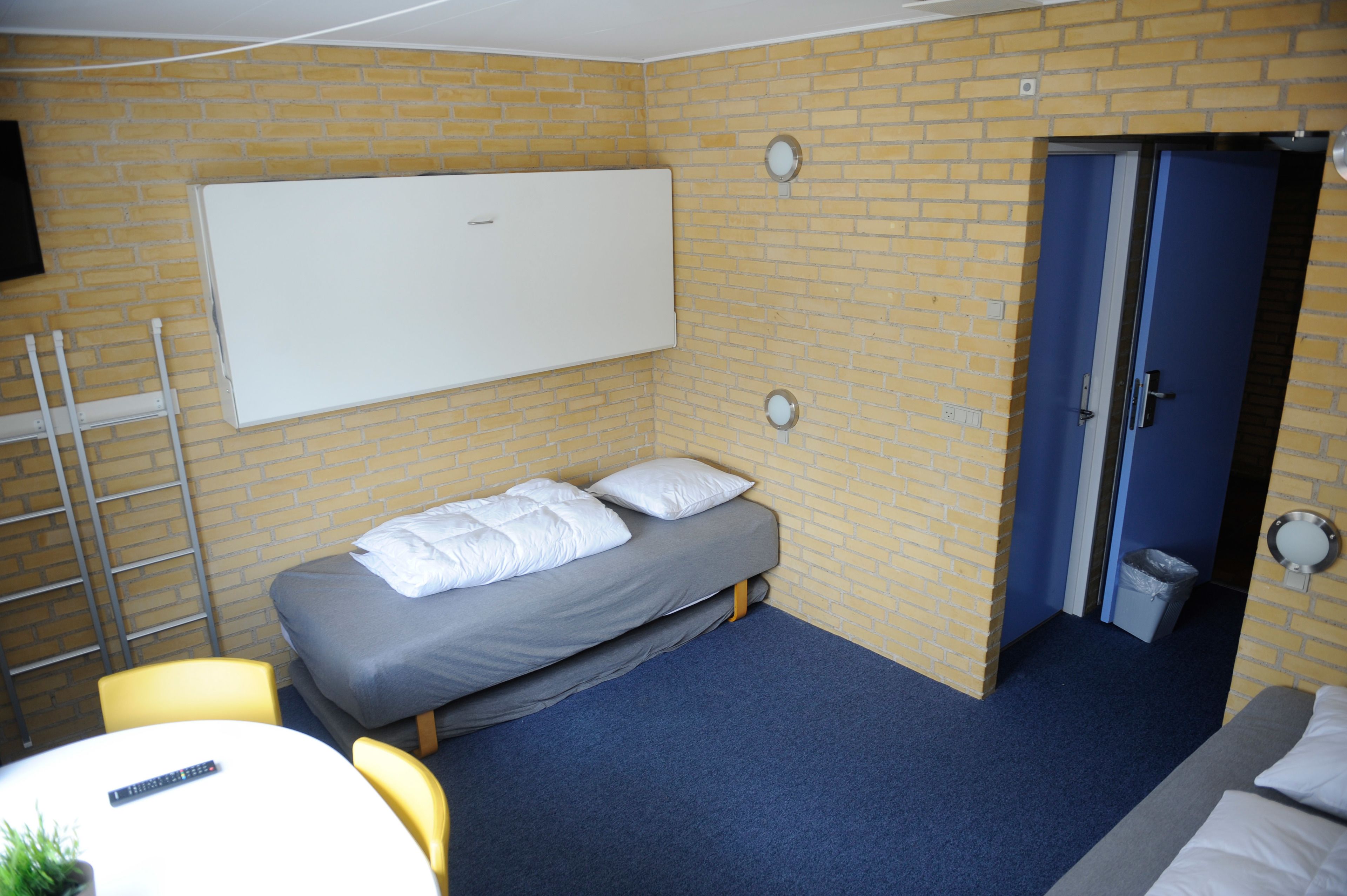 Quarto compacto com cama, uma mesa pequena, uma cadeira, um quadro branco e uma porta.