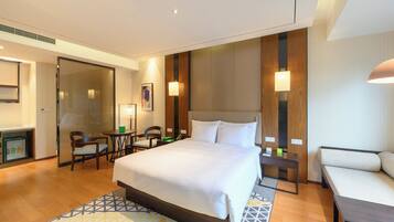 Quarto de hotel com cama, mesas de cabeceira, escrivaninha, cadeira e um pequeno frigorífico.