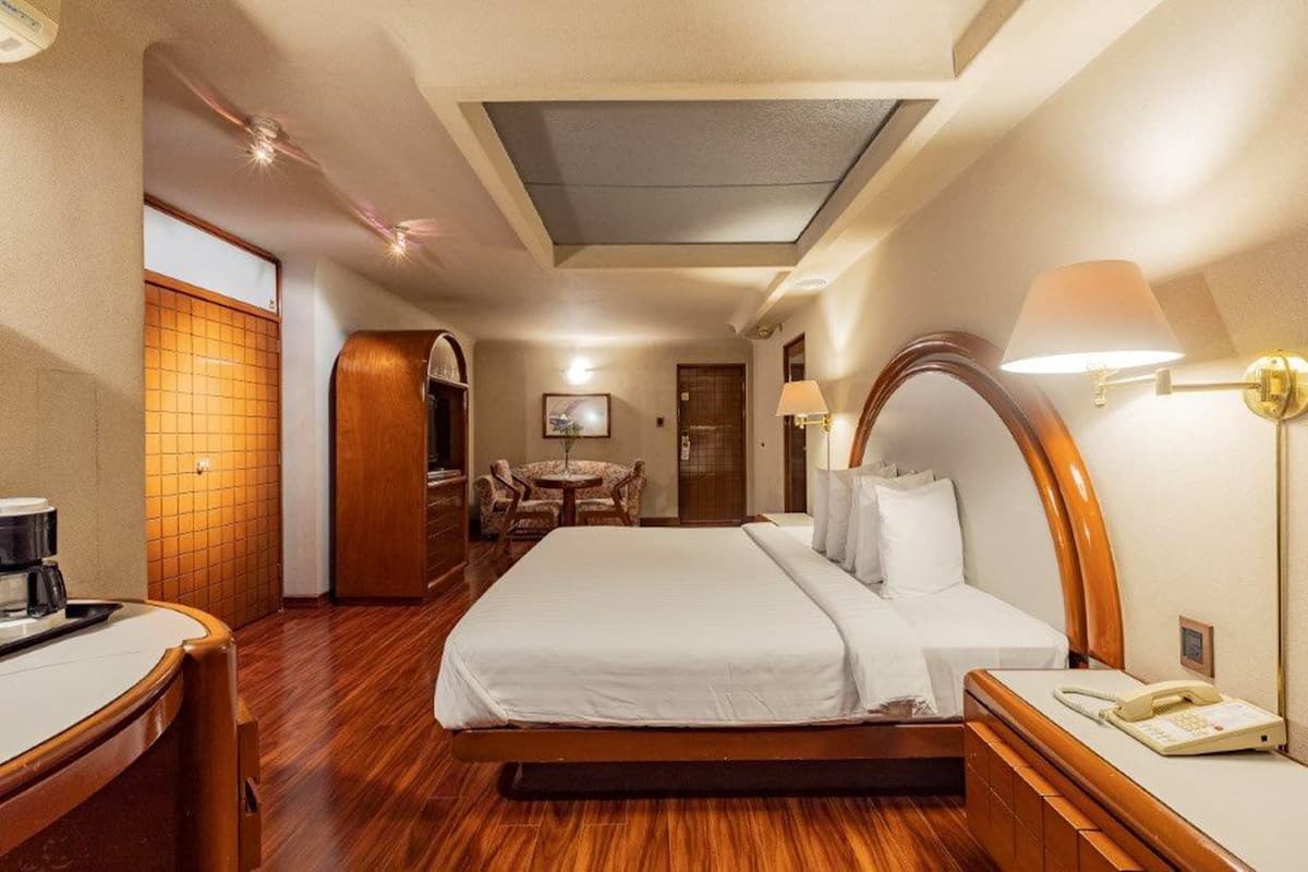 Quarto de hotel com uma cama grande, mesa de cabeceira, abajur e escrivaninha com telefone.