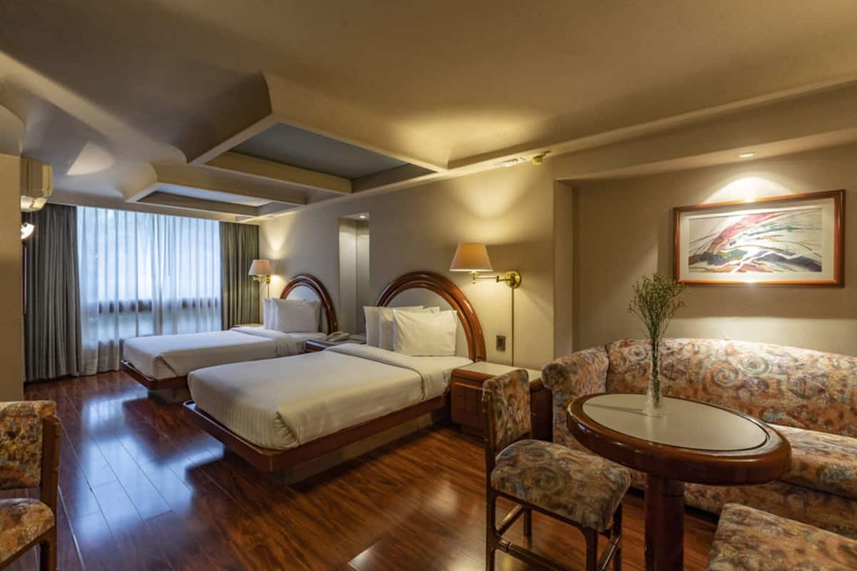Quarto de hotel com duas camas, um sofá, uma mesinha e um abajur.