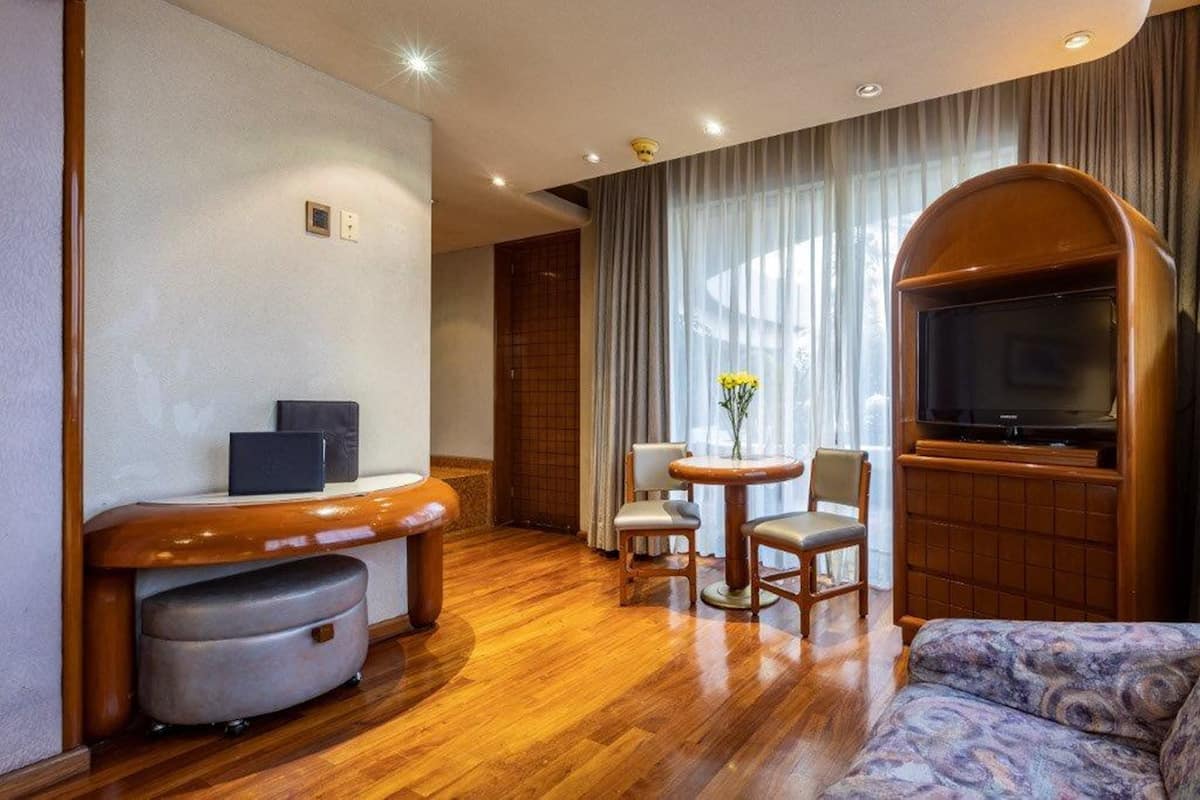 Quarto de hotel com piso de madeira, sofá, televisão, mesa pequena com cadeiras e janela com cortinas.